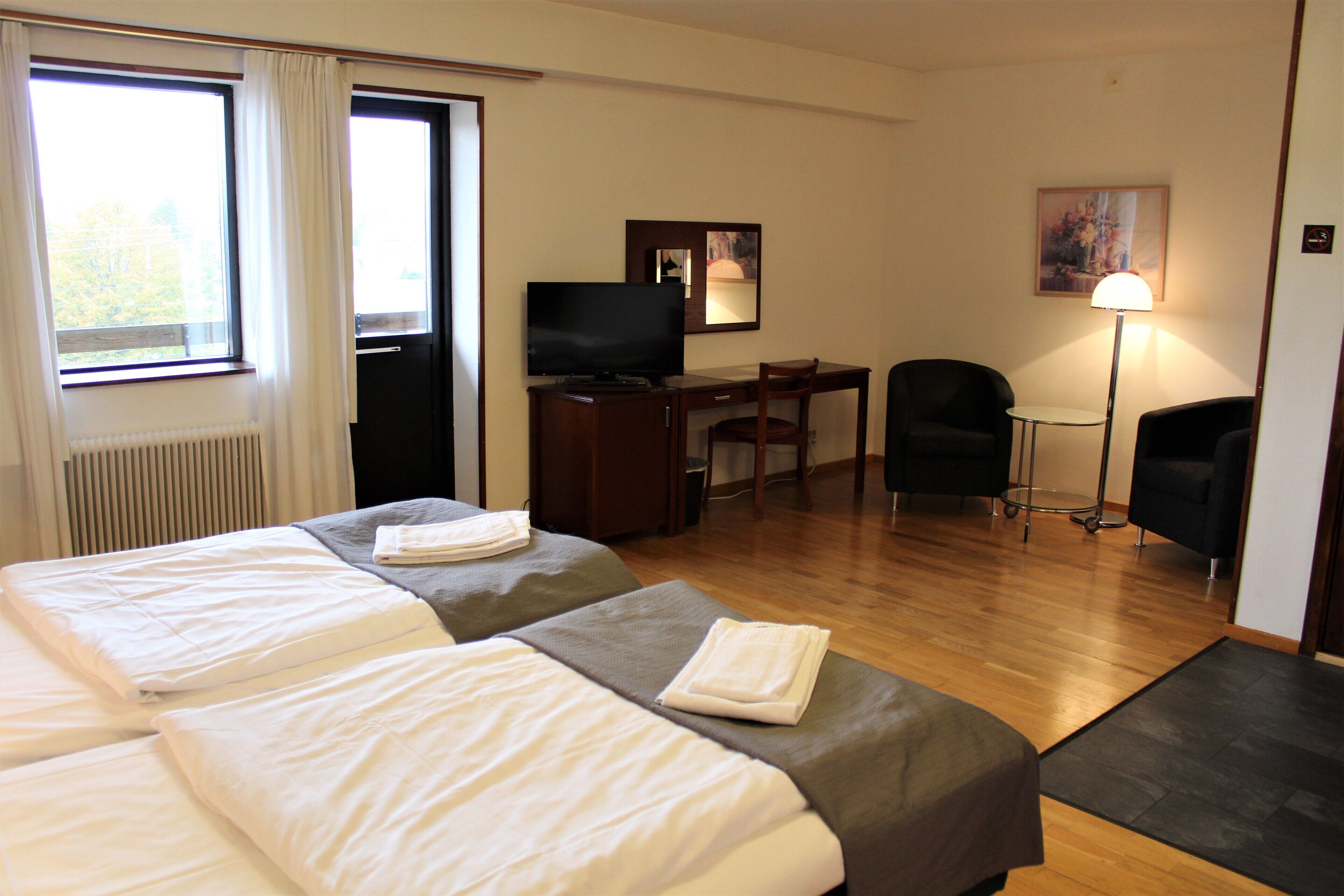 Quarto de hotel com duas camas, uma escrivaninha, uma televisão, uma cadeira e um abajur.