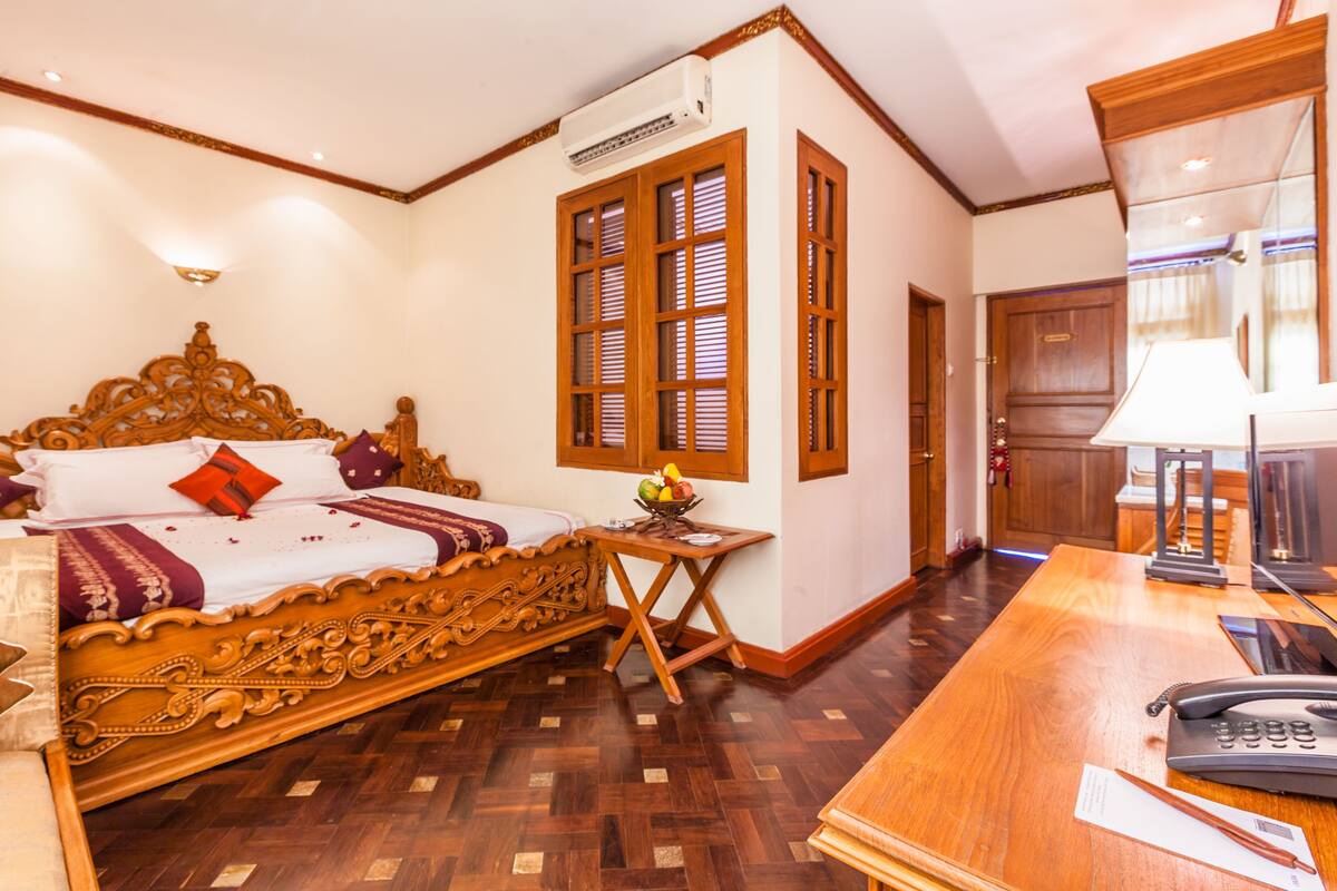Quarto de hotel com cama de madeira, uma pequena mesa com abajur, telefone e ar condicionado fixado na parede.