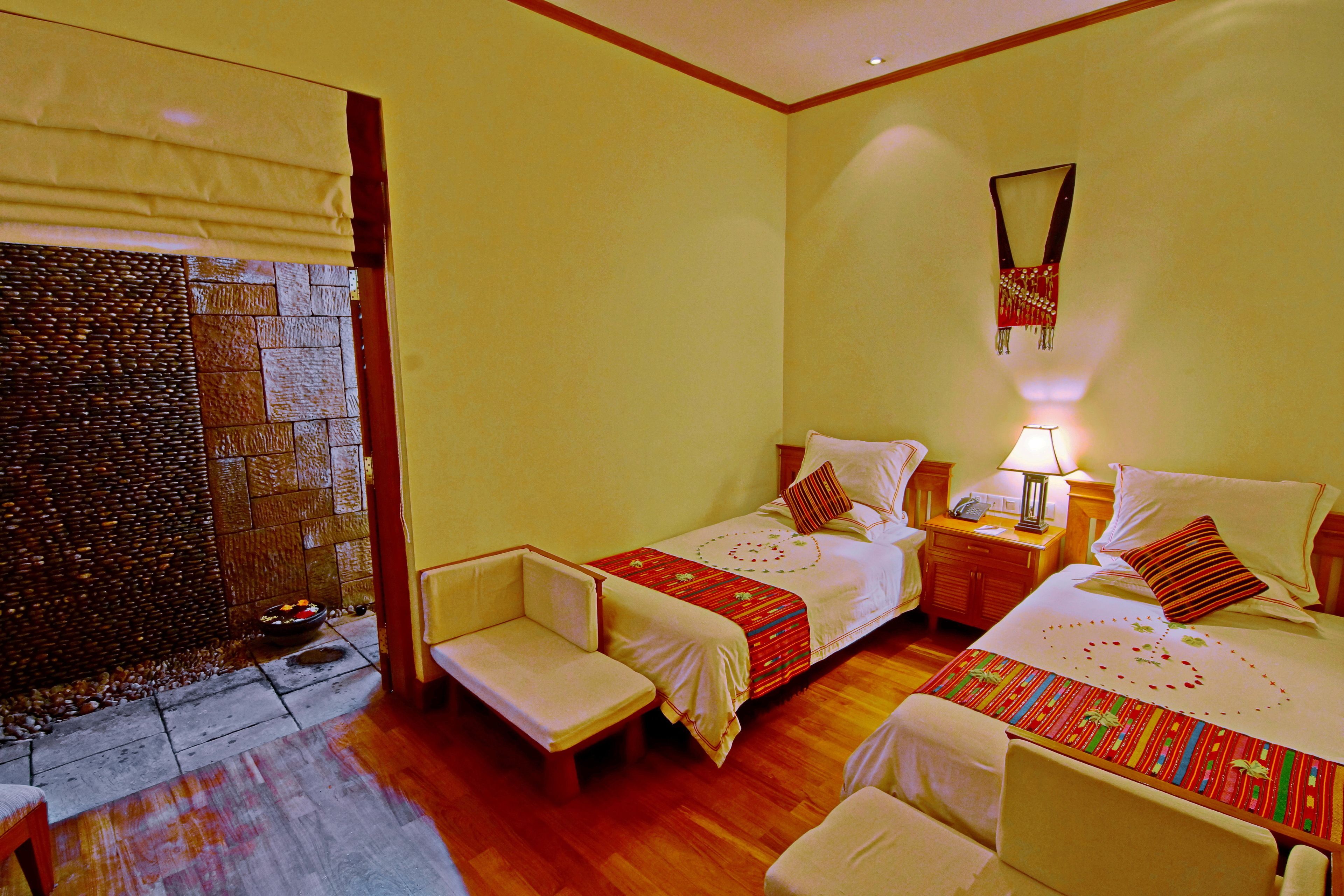 Quarto de hotel com duas camas, um espaço de estar pequeno, uma parede de pedra e uma mesa de cabeceira com luminária.