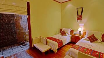 Quarto de hotel com duas camas, um espaço de estar pequeno, uma parede de pedra e uma mesa de cabeceira com luminária.