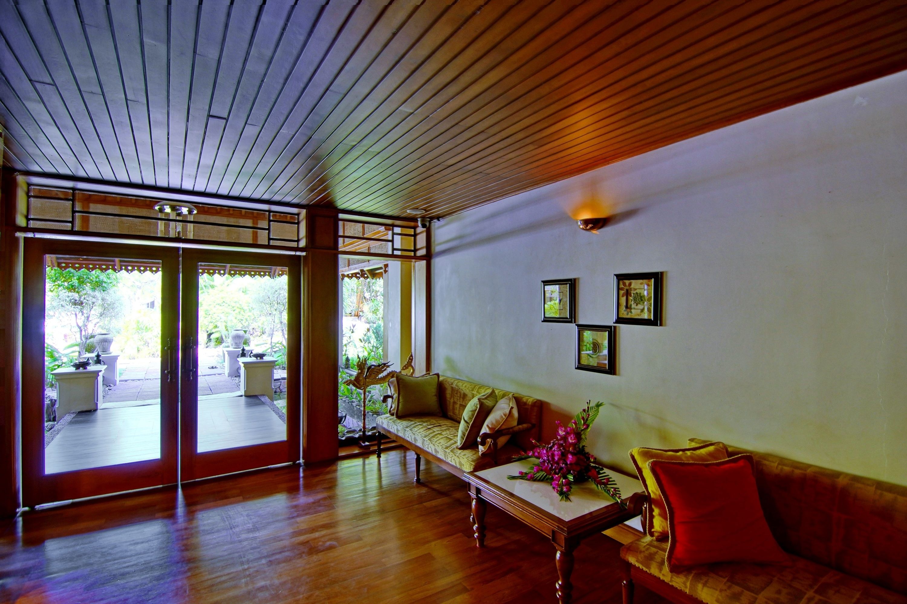 Um quarto espaçoso com teto de madeira, um sofá, uma mesa com um vaso de flores e uma porta de vidro deslizante que dá para uma área externa com plantas.