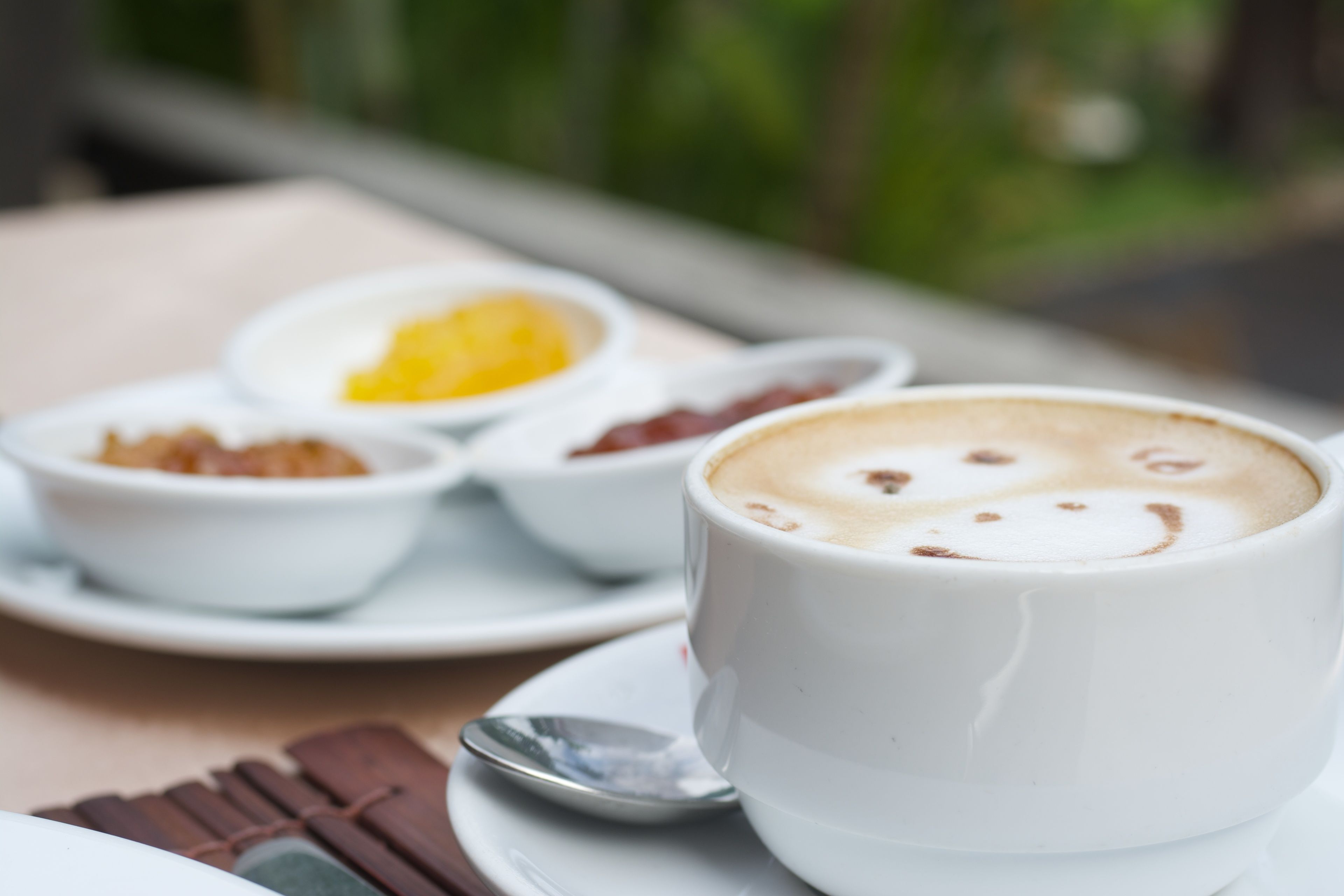Uma chávena de café com arte de latte num pires, acompanhada por pequenas taças de acompanhamentos e uma colher.