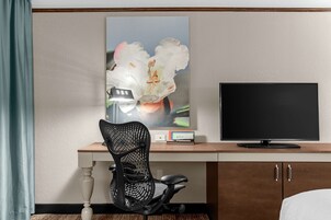 Quarto de hotel com escrivaninha de madeira, cadeira de escritório preta, televisão de tela plana e uma obra de arte floral emoldurada.