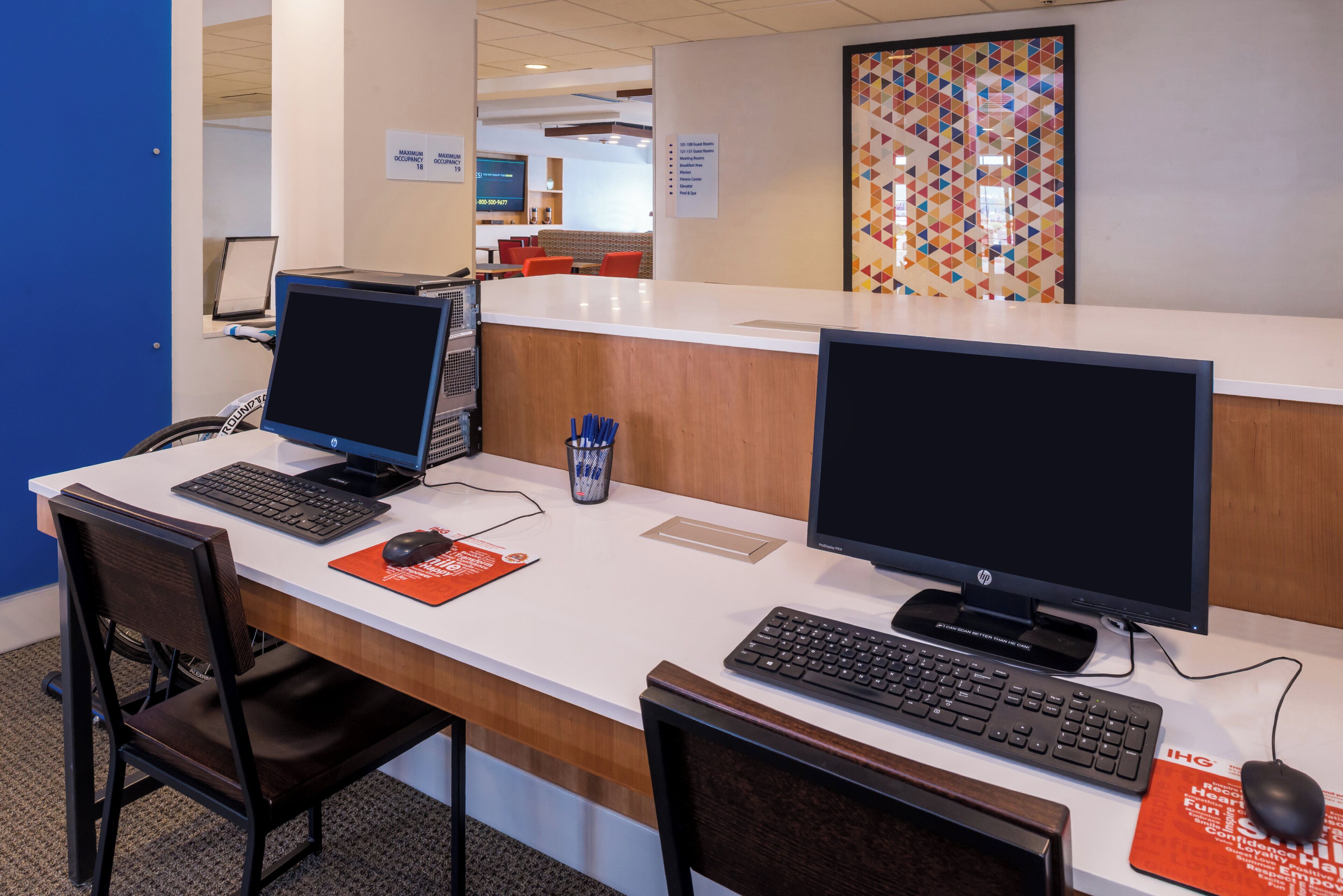 Balcão de recepção de hotel com dois monitores HP, teclado e rato sobre uma mesa branca. Há duas cadeiras em frente ao balcão e uma parede ao fundo com padrão geométrico colorido.