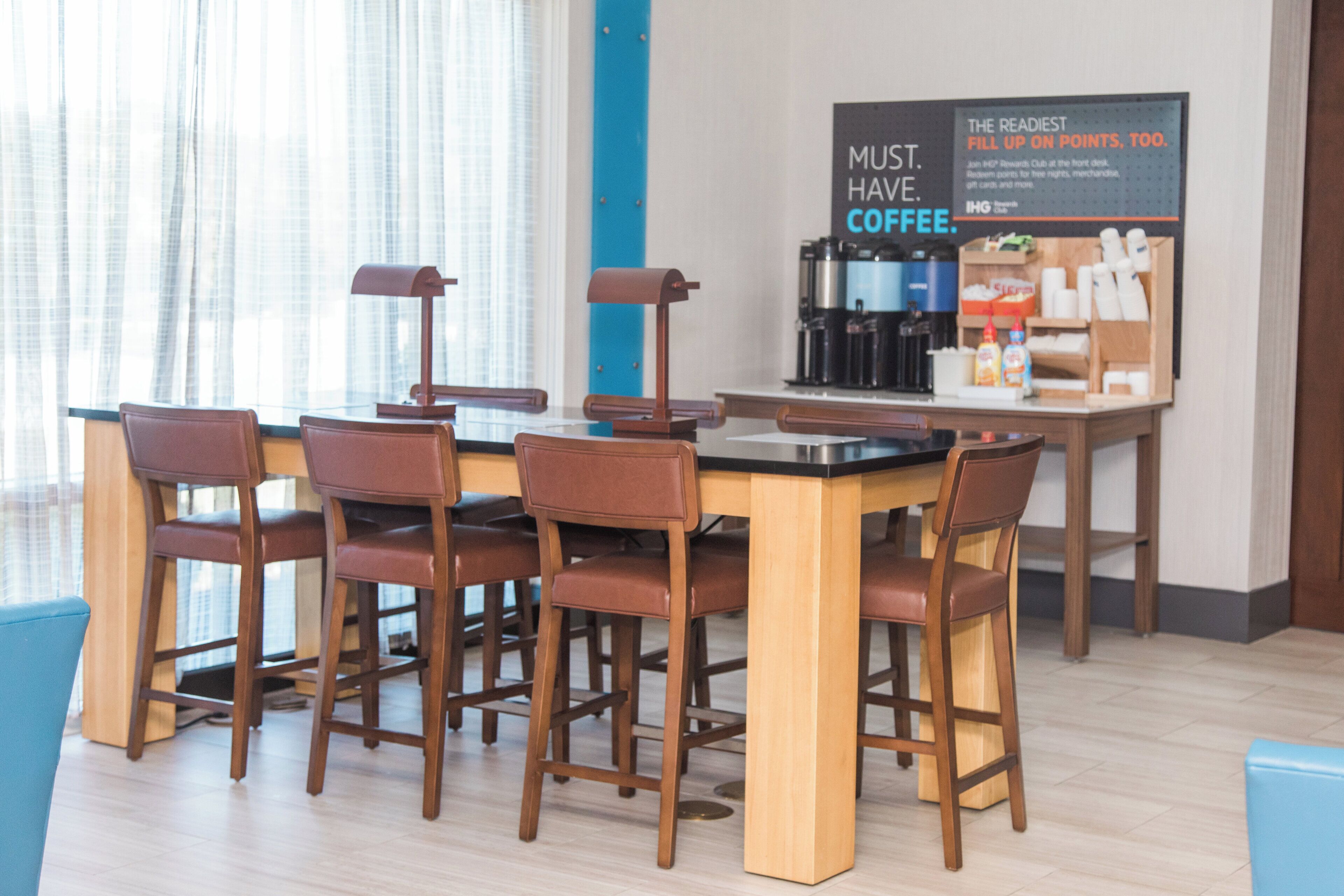 Uma área de refeições moderna com mesa e cadeiras de madeira, um espaço para café e vista da cidade através de grandes janelas.
