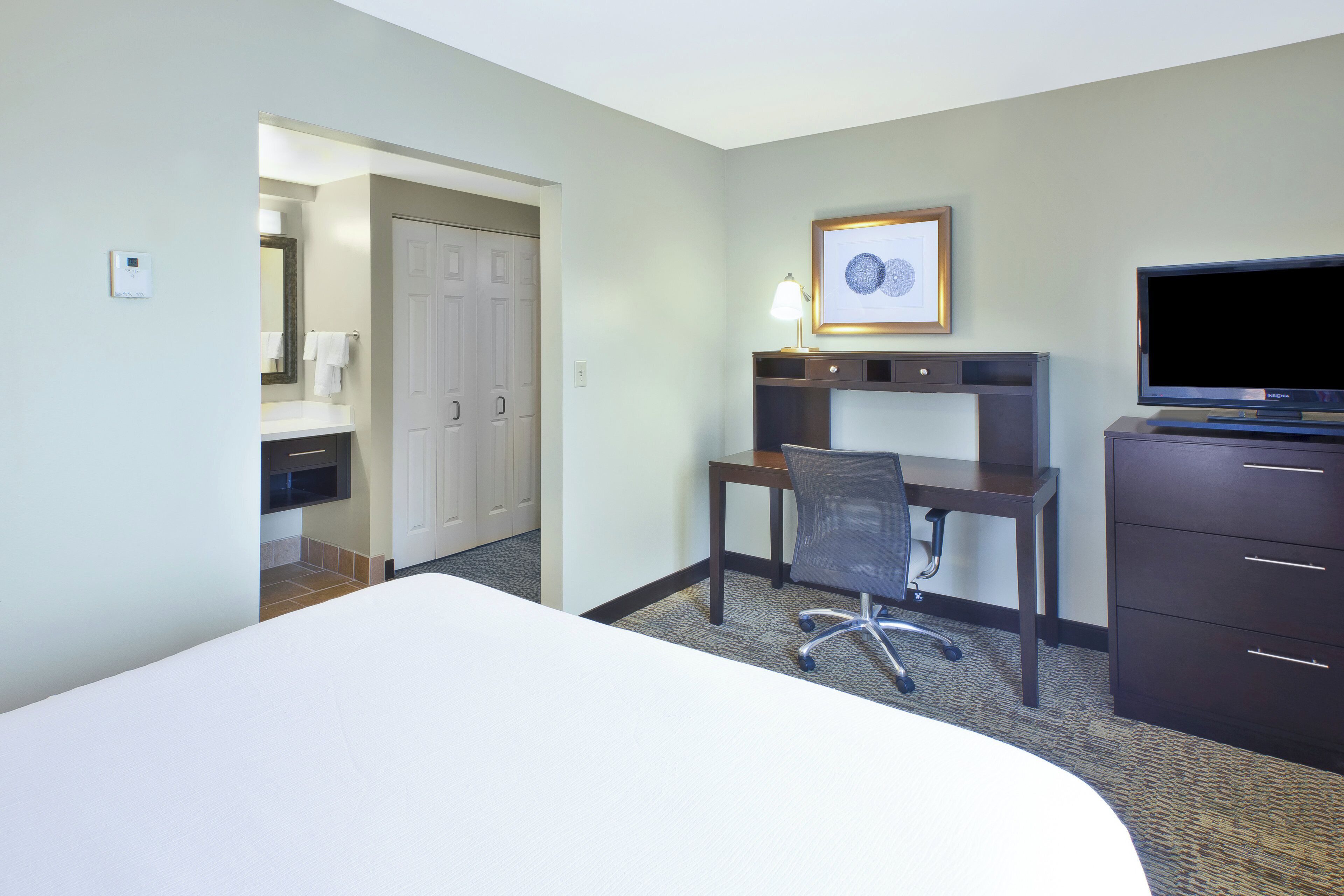 Quarto de hotel com cama, escrivaninha, cadeira, televisão e guarda-roupa.