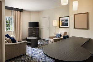 Quarto de hotel com sofá, mesa redonda, televisão, abajur e um quadro na parede.
