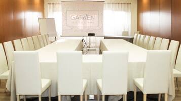 Uma sala de conferências com uma mesa comprida e cadeiras brancas, uma tela de projetor exibindo o texto "GARDEN HOTEL" e paredes de painel de madeira.