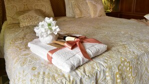 Uma cama bem arrumada com um cobre-leito estampado, um buquê de flores brancas e uma toalha dobrada com um laço rosa.