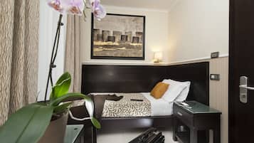 Quarto de hotel com cama, mesa de cabeceira, escrivaninha, planta em vaso e um quadro na parede.