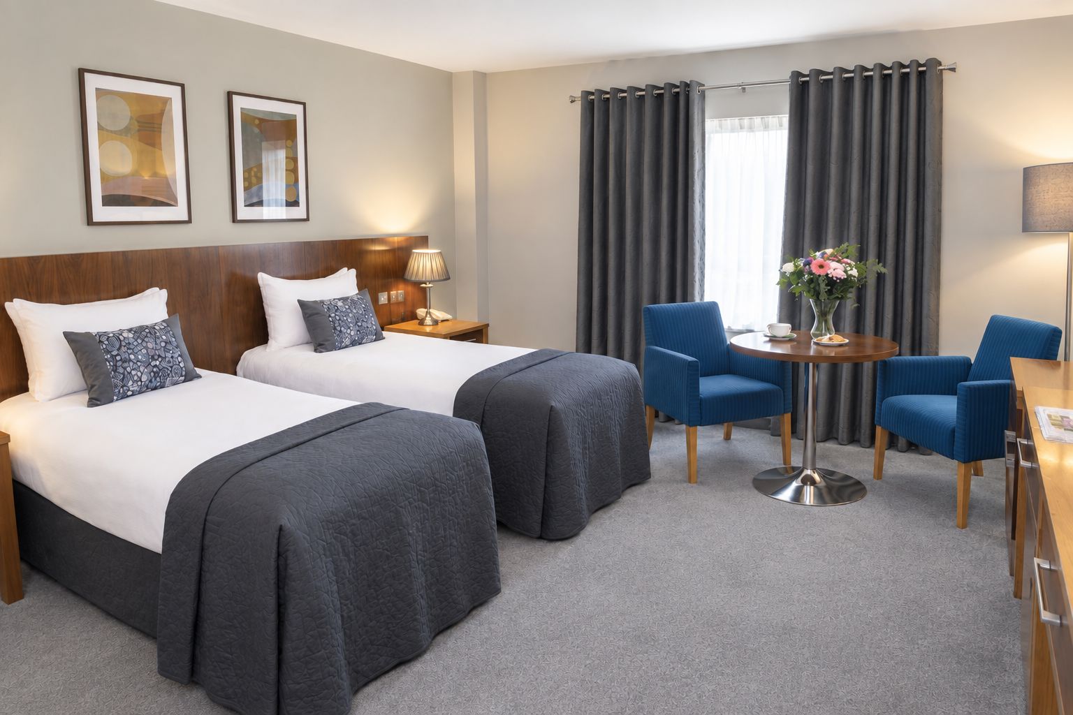 Quarto de hotel com duas camas, uma mesa pequena com cadeiras, uma luminária e uma janela com cortinas.