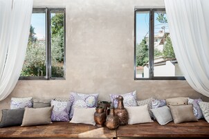 Quarto com um banco de madeira, cortinas brancas e vista para árvores e edifícios através de duas janelas.