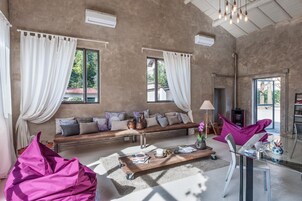 Uma sala de estar ampla, com um sofá grande, uma mesa de centro e uma mesa de jantar. Há janelas grandes com cortinas, um lustre e vista para o exterior.