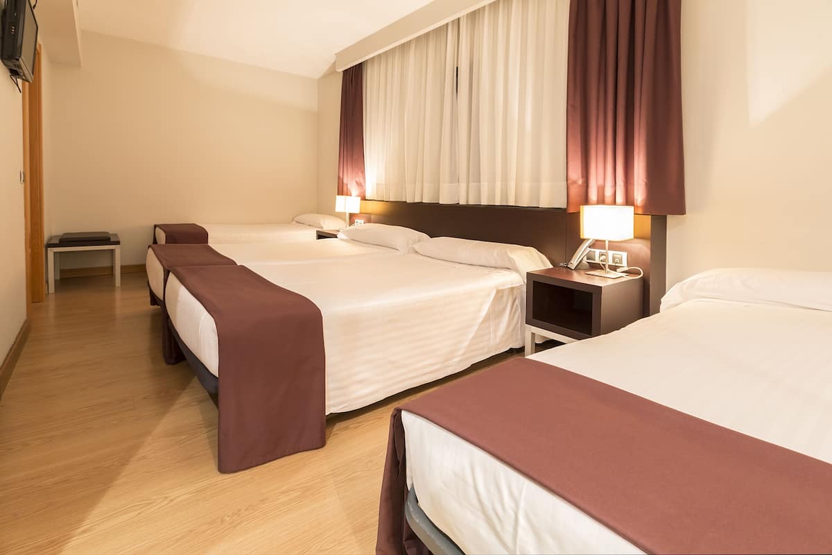 Quarto de hotel com três camas, piso de madeira, mesa de cabeceira com abajur e televisão na parede.