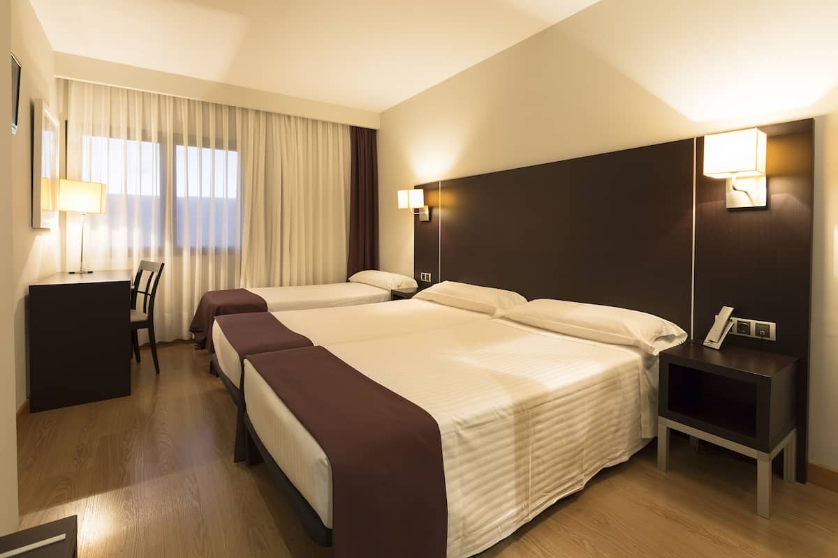 Quarto de hotel com duas camas, uma escrivaninha, uma cadeira, mesa de cabeceira com abajur e uma janela grande com cortinas.