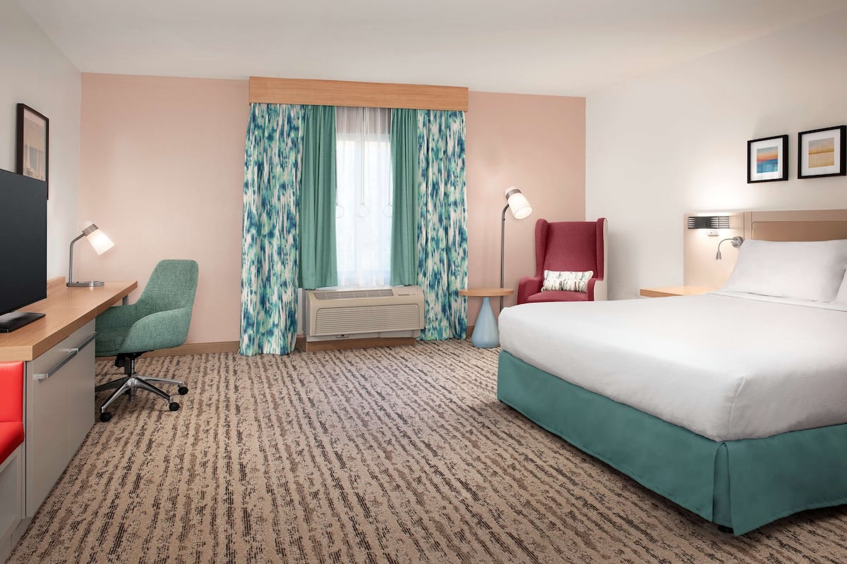 Quarto de hotel com cama, escrivaninha, cadeira e janela com cortinas.