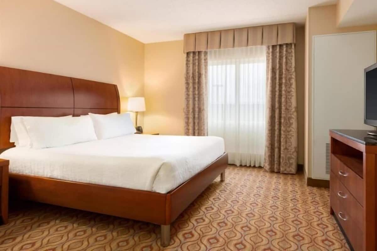 Quarto de hotel com uma cama, televisão, penteadeira e janela com cortinas.
