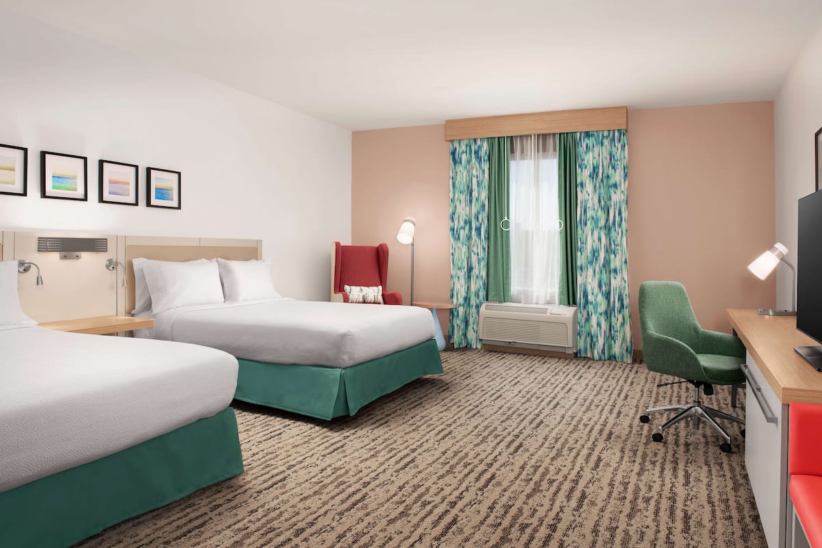 Quarto de hotel com duas camas, uma escrivaninha com cadeira, televisão e janela com cortinas.