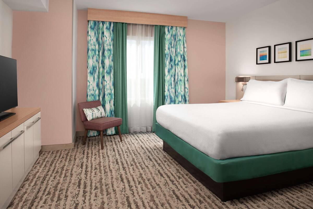 Quarto de hotel com uma cama grande, uma mesa de trabalho com televisão, uma cadeira e uma janela com cortinas.