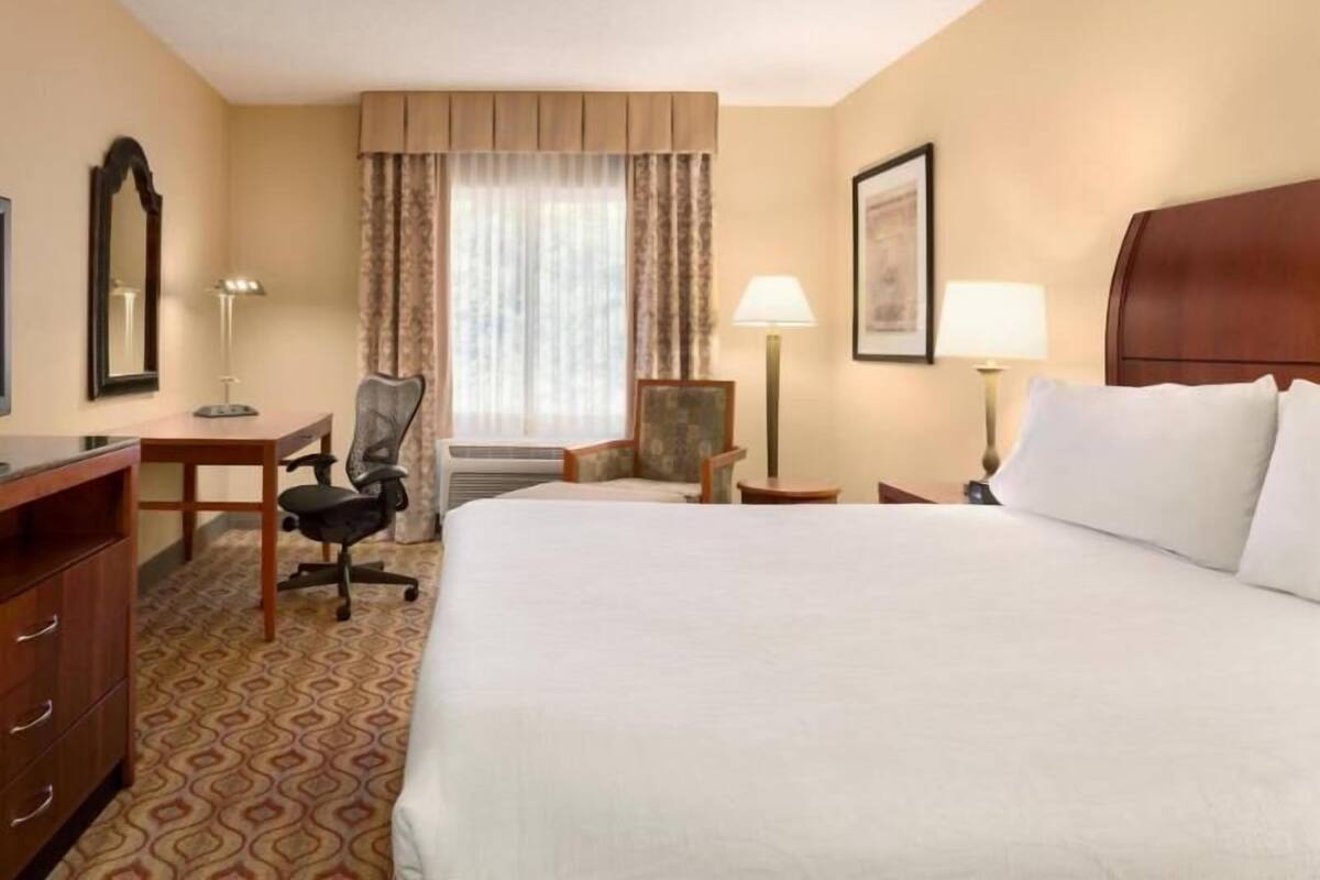 Quarto de hotel com cama, escrivaninha, cadeira, luminárias e janela com cortinas.