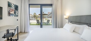 Quarto de hotel com uma cama grande, uma mesa e uma cadeira. Há uma varanda com vista para a praia e palmeiras.