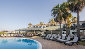 Área da piscina com espreguiçadeiras e guarda-sóis, céu claro e um edifício com varandas.