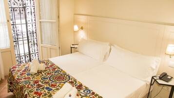 Quarto de hotel com uma cama grande, mesa de cabeceira com abajur e janela com grade de ferro decorativa.