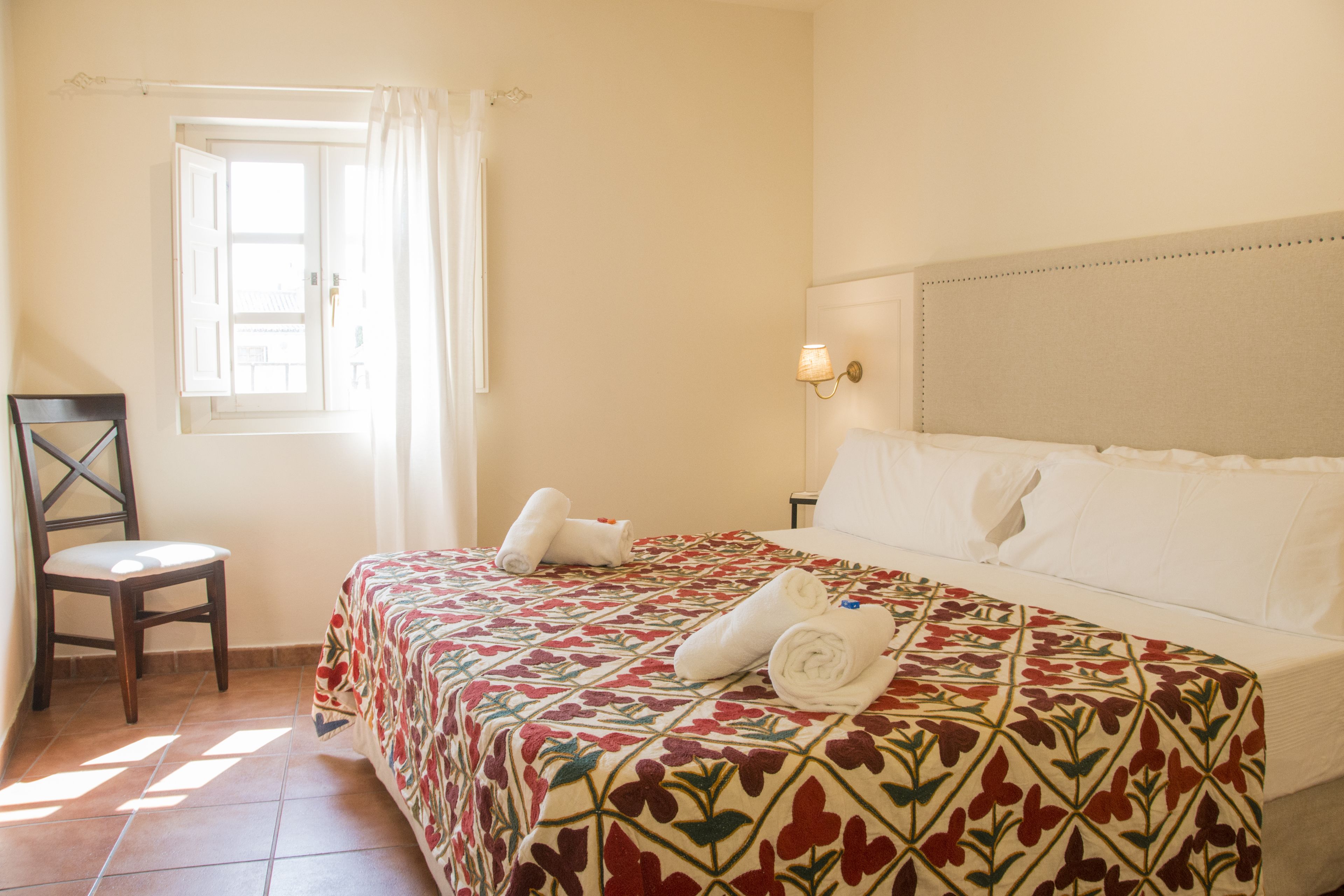 Quarto de hotel com cama, cadeira, janela com cortinas e mesa de cabeceira com abajur.