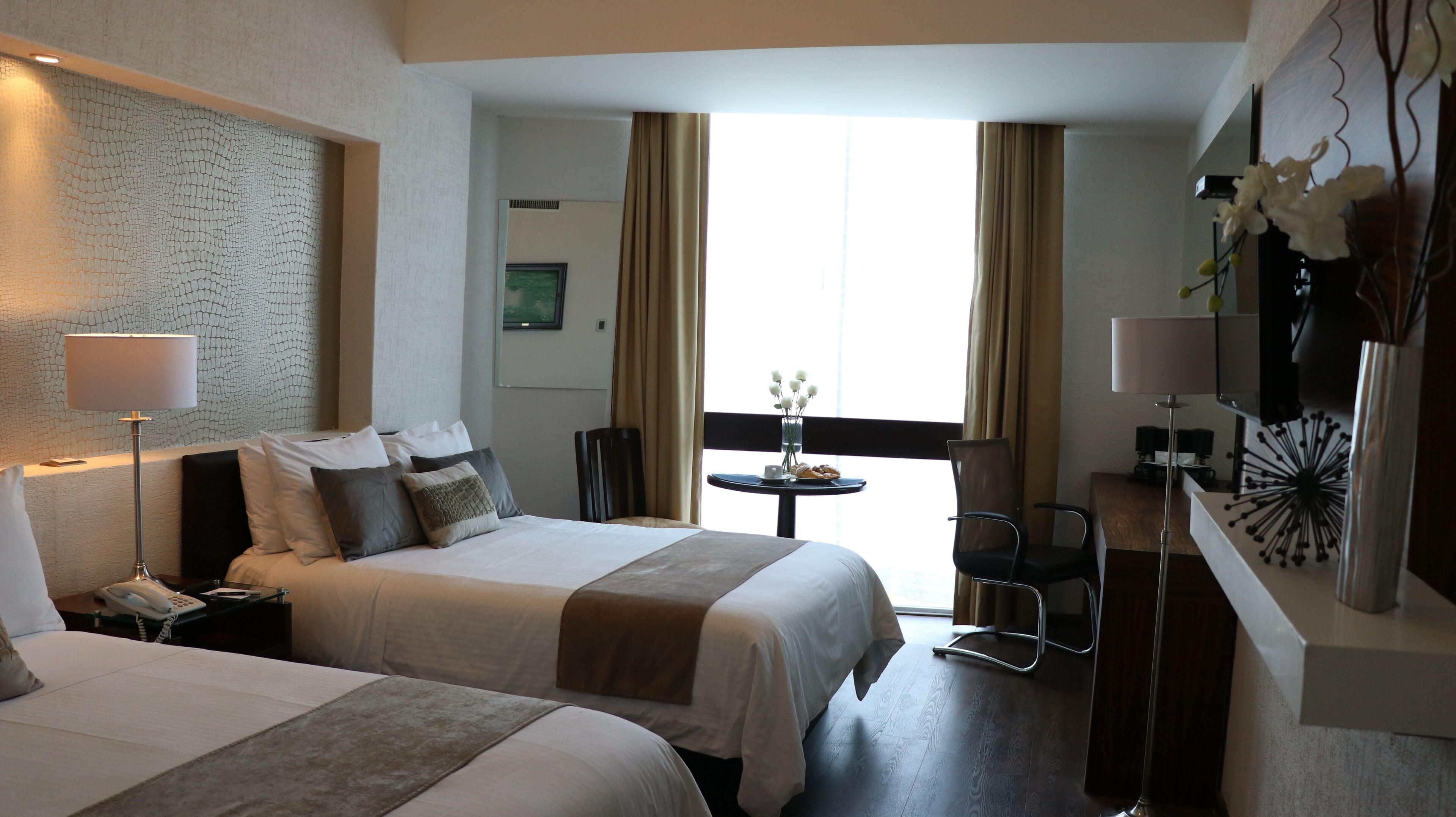 Quarto de hotel com duas camas, uma mesa, uma cadeira, um abajur, um telefone, um vaso com flores e uma janela grande com cortinas.