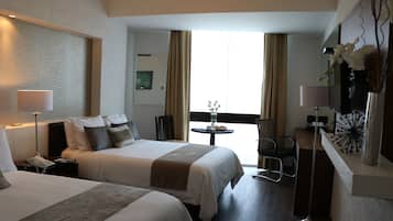 Quarto de hotel com duas camas, uma mesa, uma cadeira, um abajur, um telefone, um vaso com flores e uma janela grande com cortinas.
