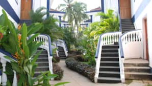 Um caminho leva a um edifício com detalhes em branco e azul, uma escadaria e vegetação exuberante, incluindo palmeiras.