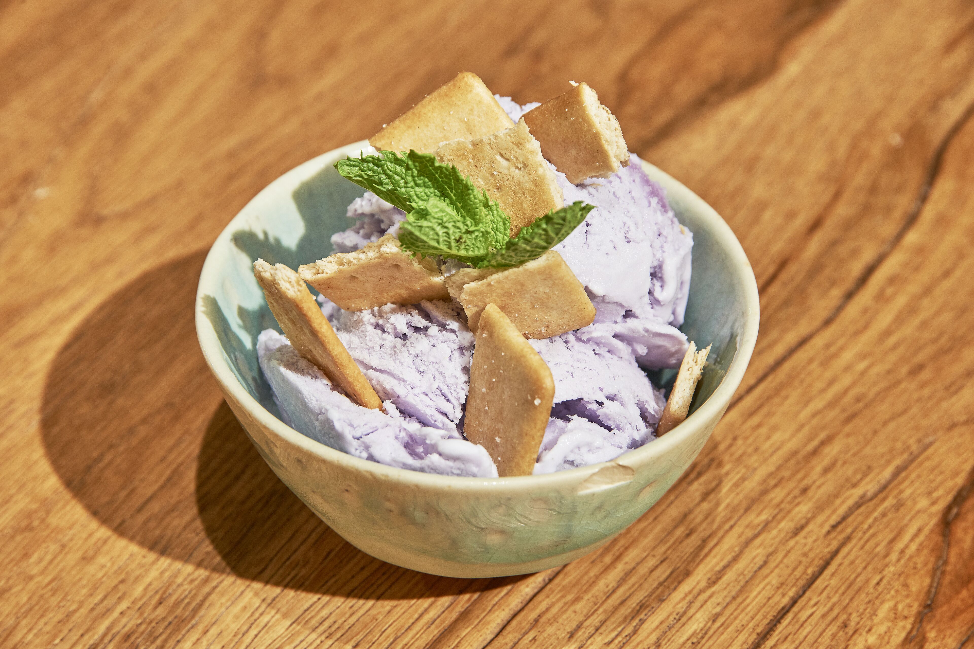 Uma taça de sorvete roxo com pedaços de biscoito e uma folha de hortelã sobre uma mesa de madeira.