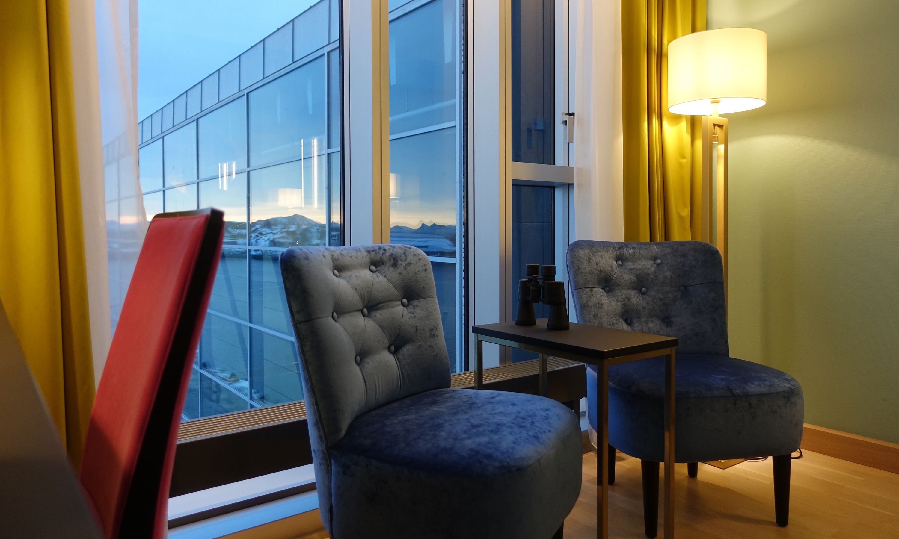 Quarto moderno com duas poltronas estofadas, uma pequena mesa e vista para a paisagem urbana através de amplas janelas.