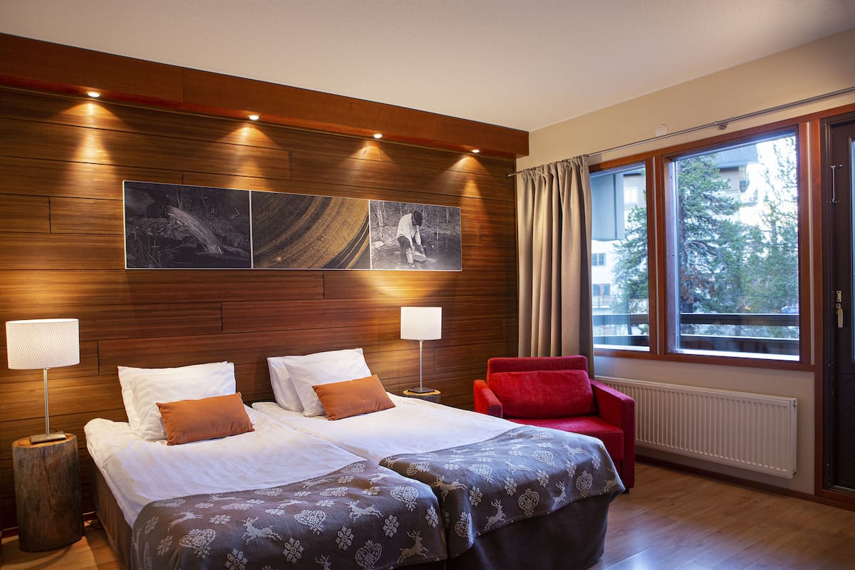 Quarto de hotel com cabeceira de madeira, duas camas, uma poltrona vermelha e uma janela grande com vista para edifícios.