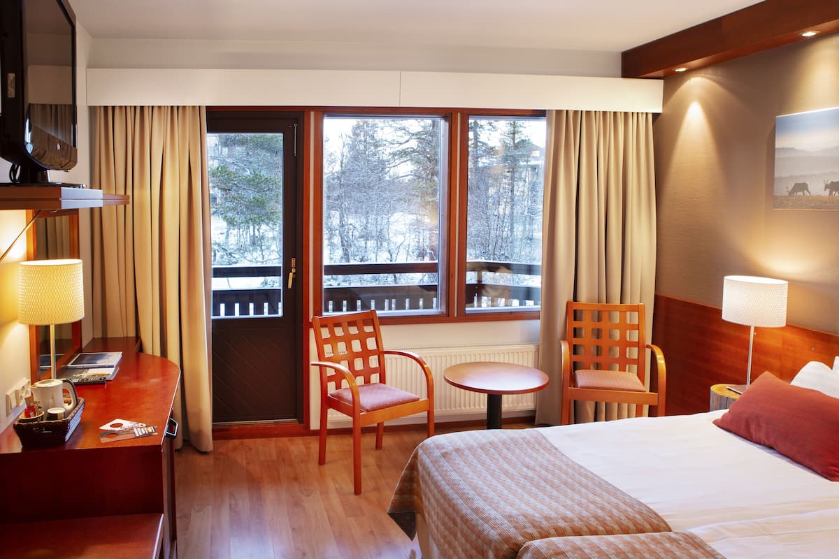 Quarto de hotel com uma cama, duas cadeiras, uma mesa pequena, uma escrivaninha com abajur e uma janela com cortinas.