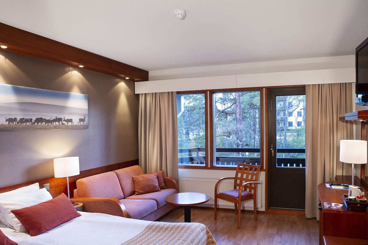 Quarto de hotel com uma cama, um sofá, uma poltrona, uma mesa, um abajur, um quadro e uma janela com cortinas.