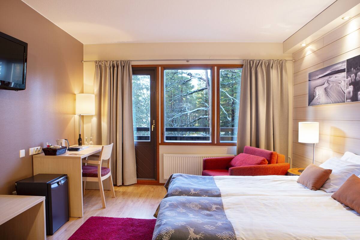 Quarto de hotel com cama, escrivaninha, cadeira e vista para árvores pela janela.