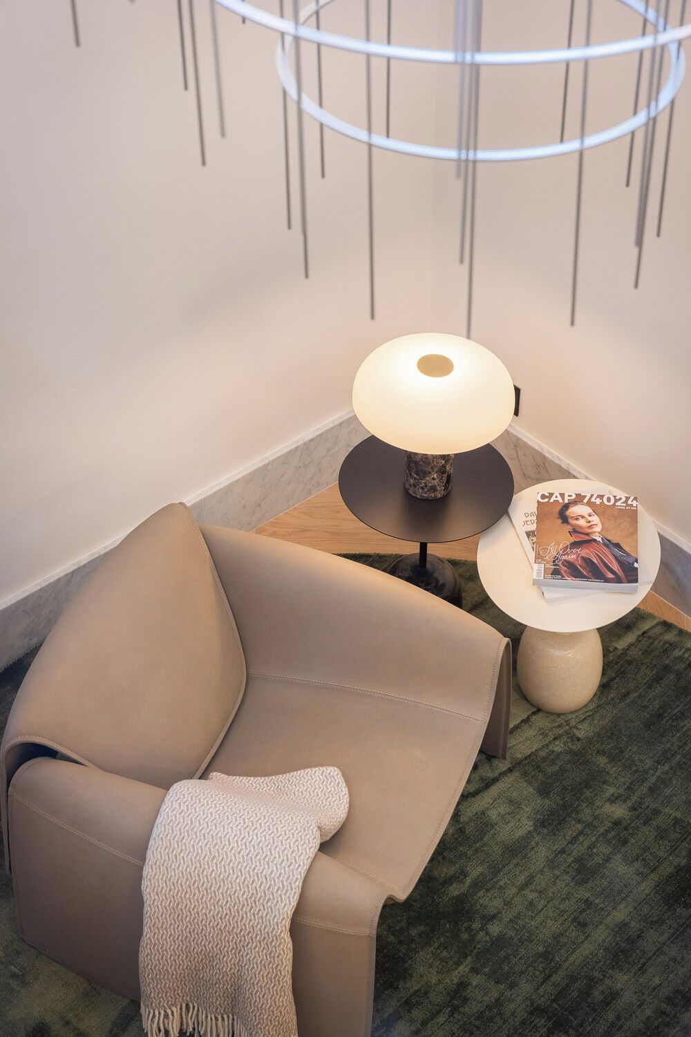 Uma sala de estar moderna com um sofá bege, uma mesa lateral redonda com abajur e uma revista.