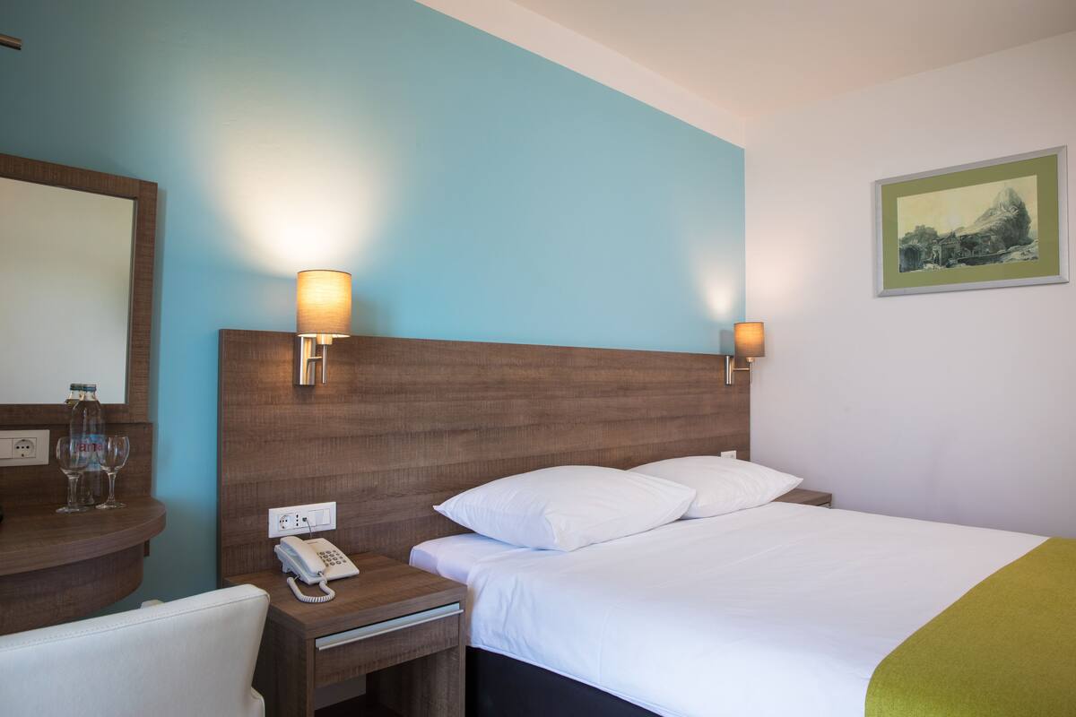 Quarto de hotel com cabeceira de madeira, cama com roupa de cama branca, mesa de cabeceira com luminária, telefone e espelho.