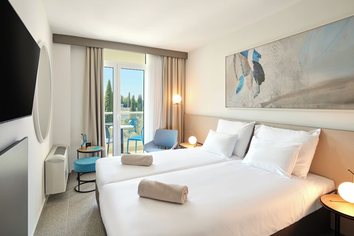 Quarto de hotel com duas camas, televisão, escrivaninha com candeeiro, cadeira e vista para árvores pela janela.