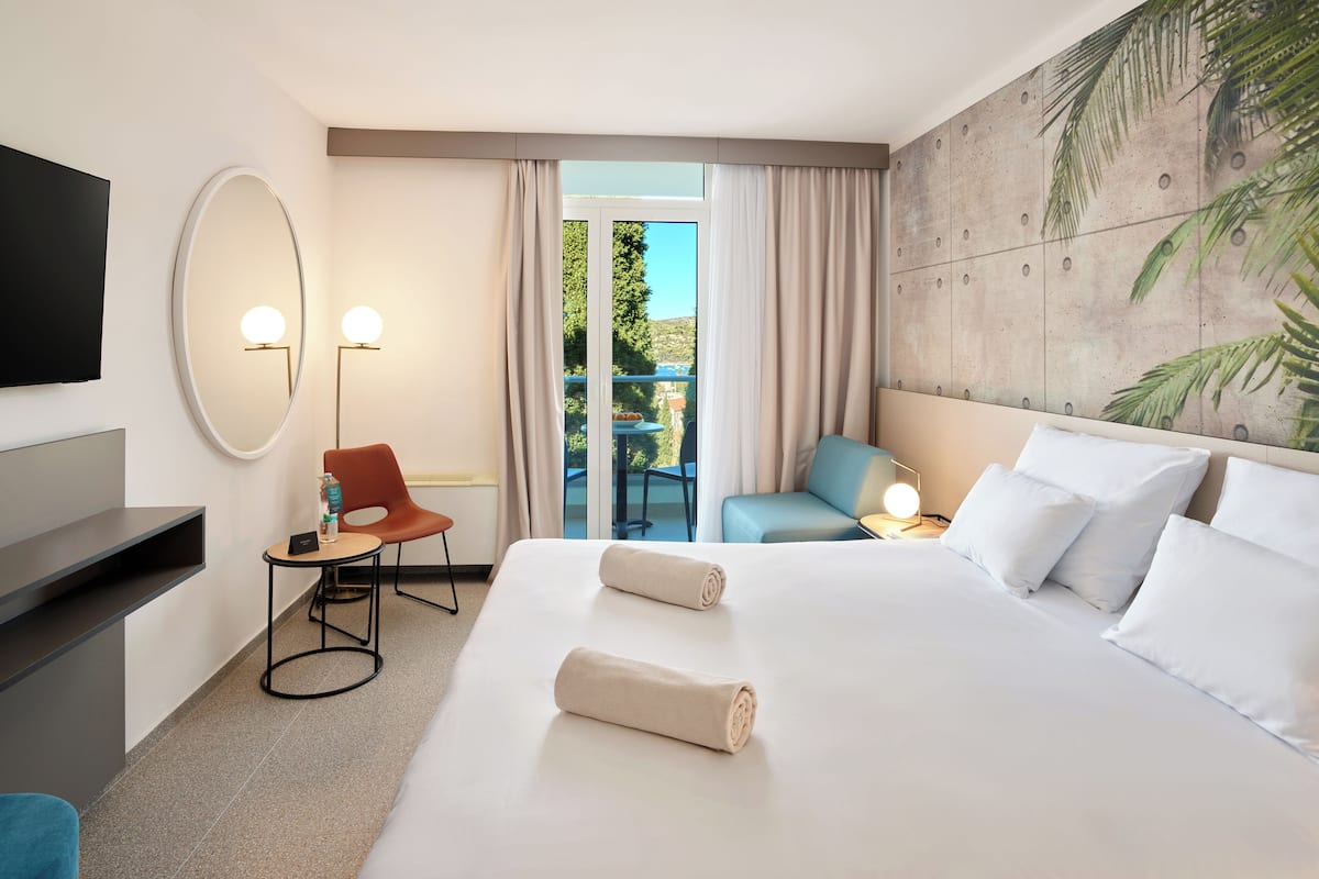 Quarto de hotel com cama, poltrona, escrivaninha, televisão e varanda com vista.