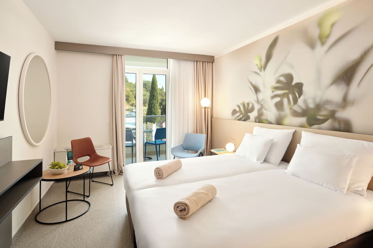 Quarto de hotel com duas camas, uma escrivaninha com cadeira, televisão e varanda com vista para árvores e um edifício.