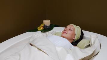 Uma pessoa deitada em uma cama com cobertor branco, travesseiro verde e uma vela sobre uma folha verde.