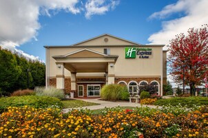 Edifício Holiday Inn Express com entrada coberta, jardim com plantas e flores coloridas e céu azul claro.