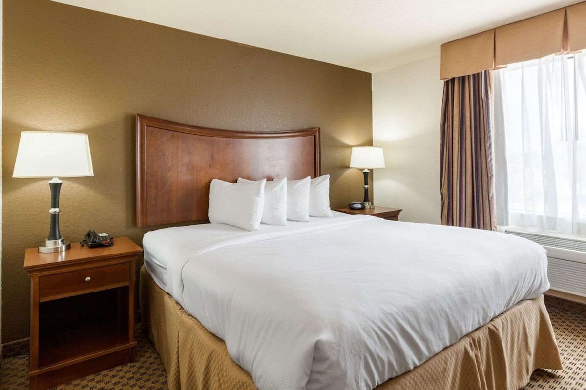 Quarto de hotel com uma cama grande, duas mesas de cabeceira com luminárias, uma cabeceira de madeira e uma janela com cortinas.