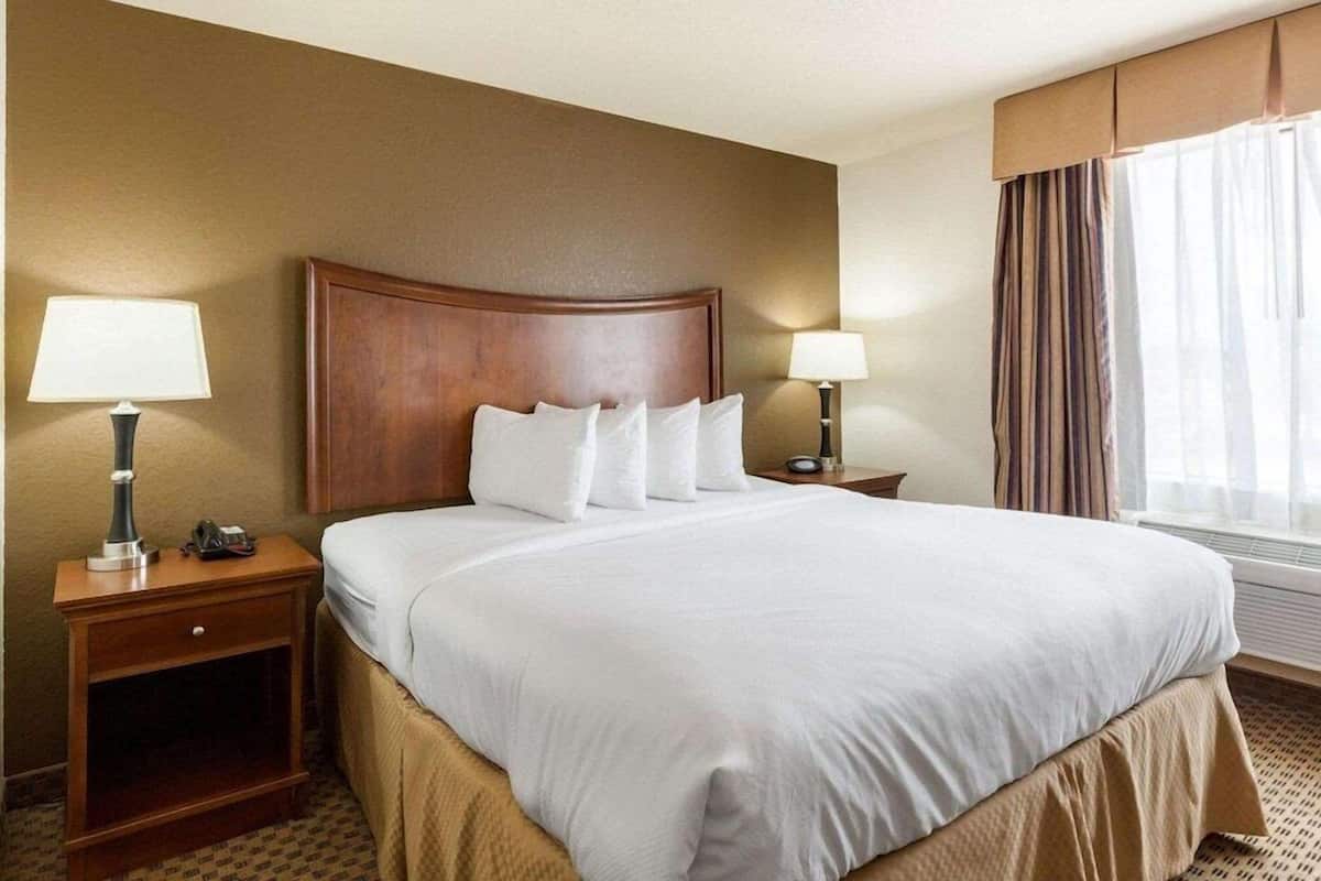 Quarto de hotel com uma cama grande, duas mesas de cabeceira com luminárias, uma cabeceira de madeira e uma janela com cortinas.
