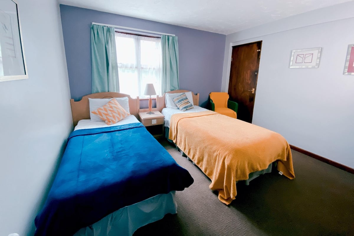 Quarto com duas camas, janela com cortinas, uma cadeira e uma porta.