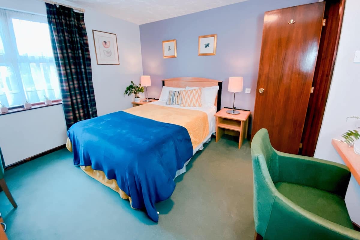 Quarto de hotel com uma cama, duas mesas de cabeceira, uma poltrona verde, uma porta de madeira e uma janela com cortinas.