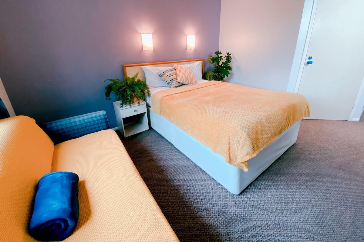 Quarto de hotel com uma cama, duas mesas de cabeceira, um criado-mudo, uma planta em vaso e uma porta.
