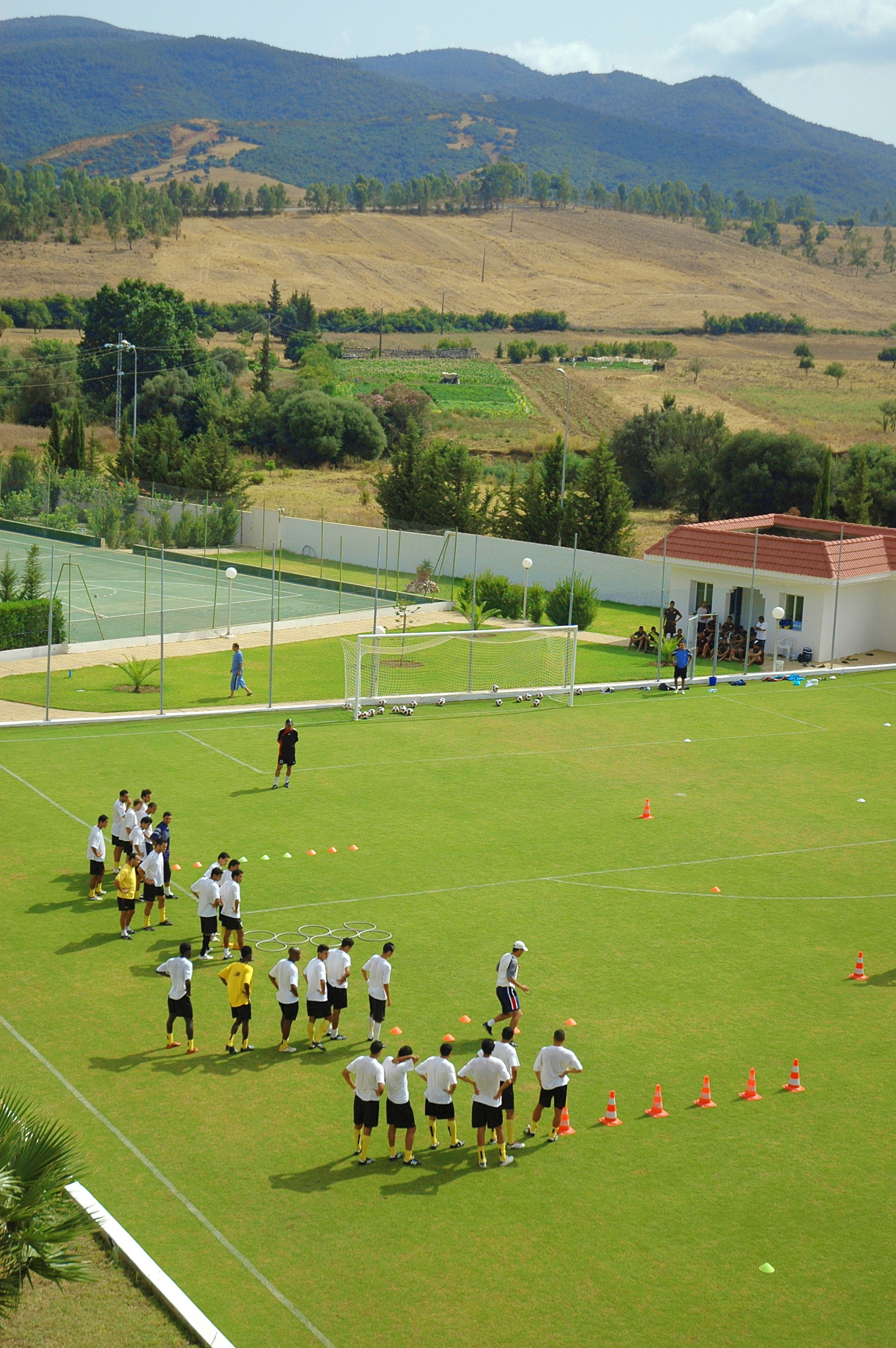 Um campo de futebol com jogadores e cones, cercado por uma grade e árvores.